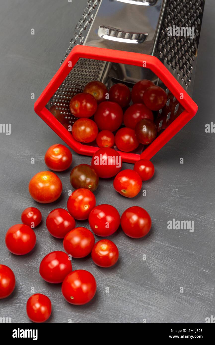 Red cherry tomatoes spill out of a red grater. Close up. Top view. Grey ...