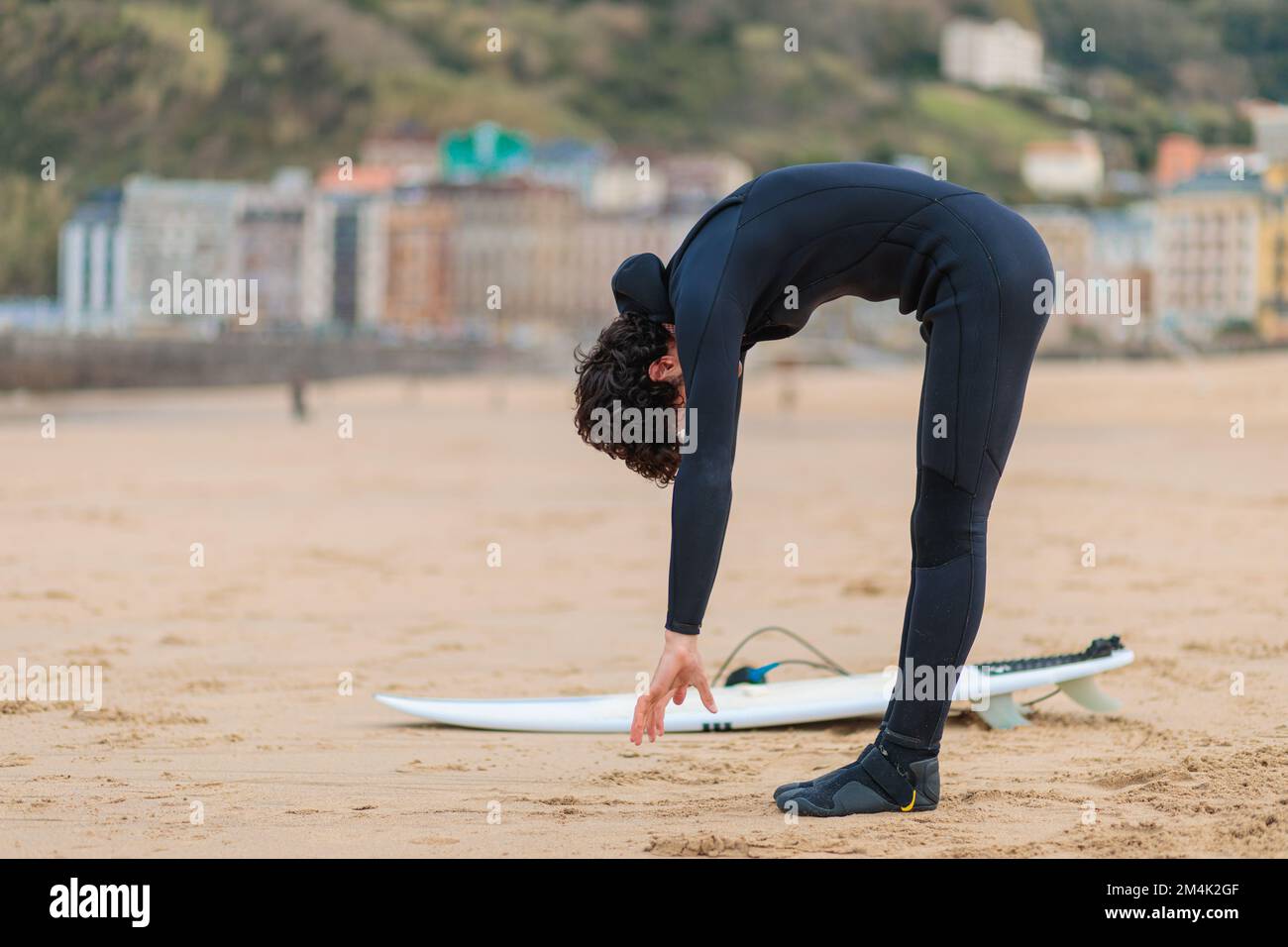 A young surfer boy in a wetsuit stretching on the beach with a surfing ...