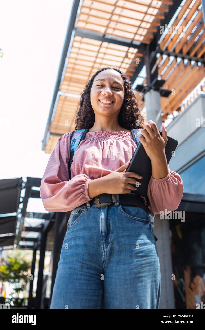 Low angle smiling latin university student woman standing and holding ...