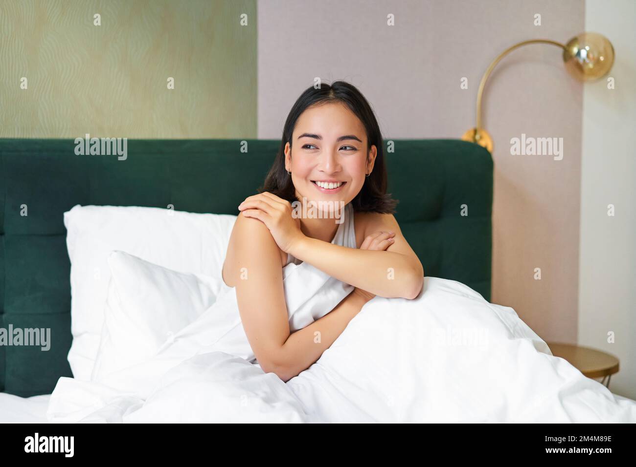 Happy mornings. Smiling asian woman wakes up in her bed, looks outside ...