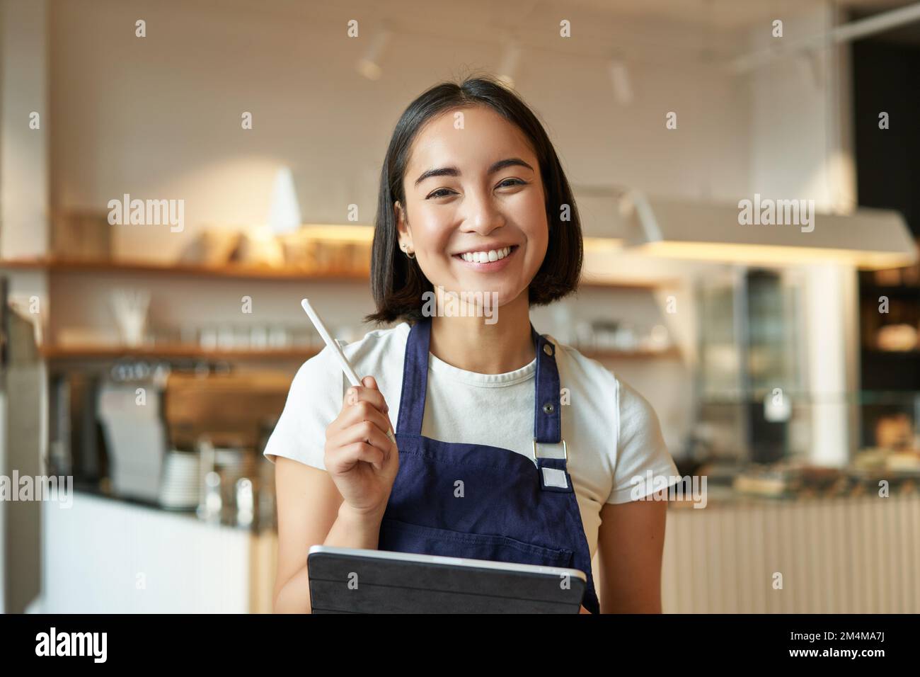 Enthusiastic asian female entrepreneur, cafe owner with tablet, wearing shop uniform, standing ...