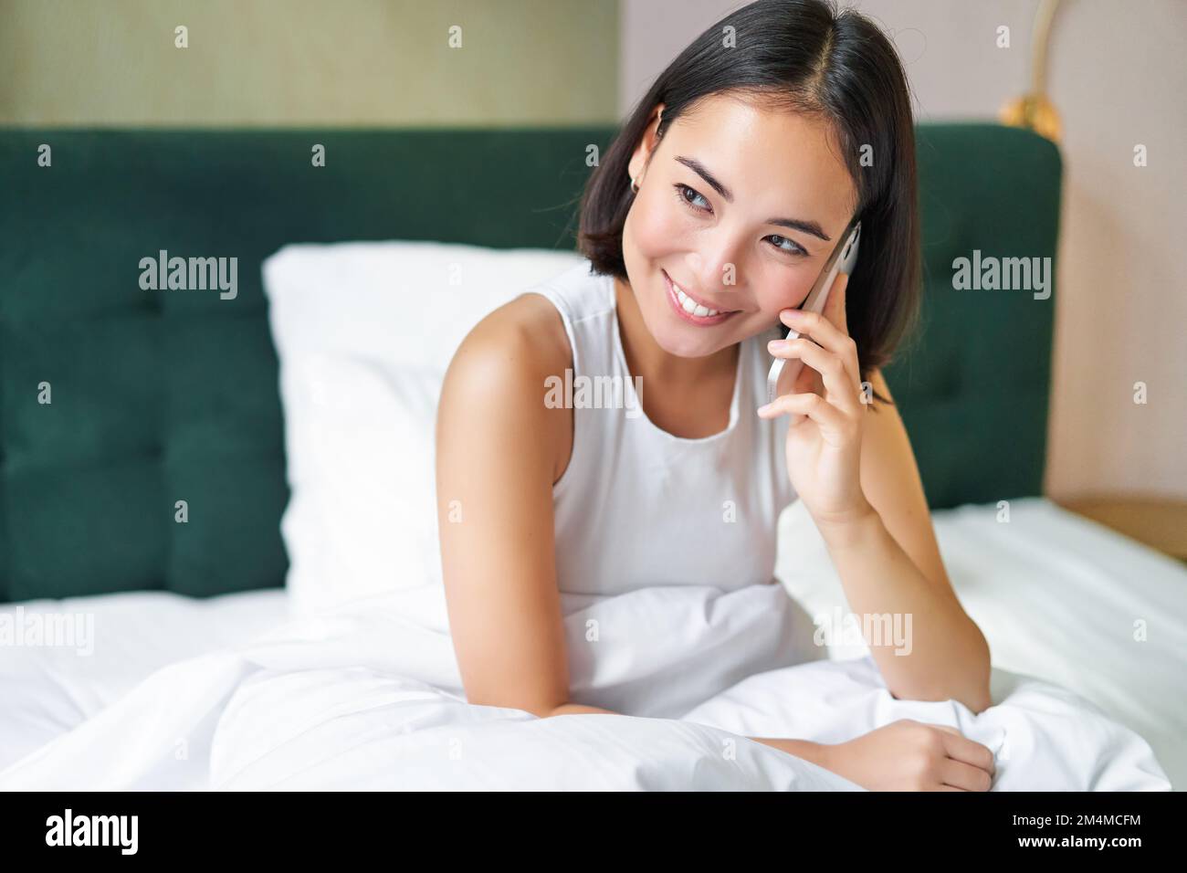 Smiling korean girl talks on mobile phone and lying in bed. Cute woman answers telephone call ...
