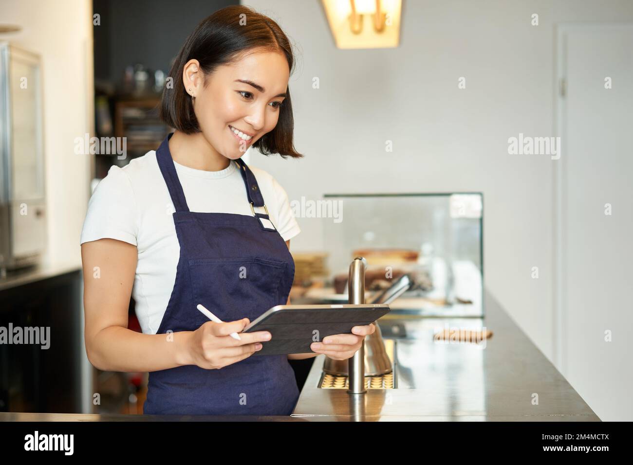 Small cafe business. Smiling asian girl barista in apron, using tablet ...