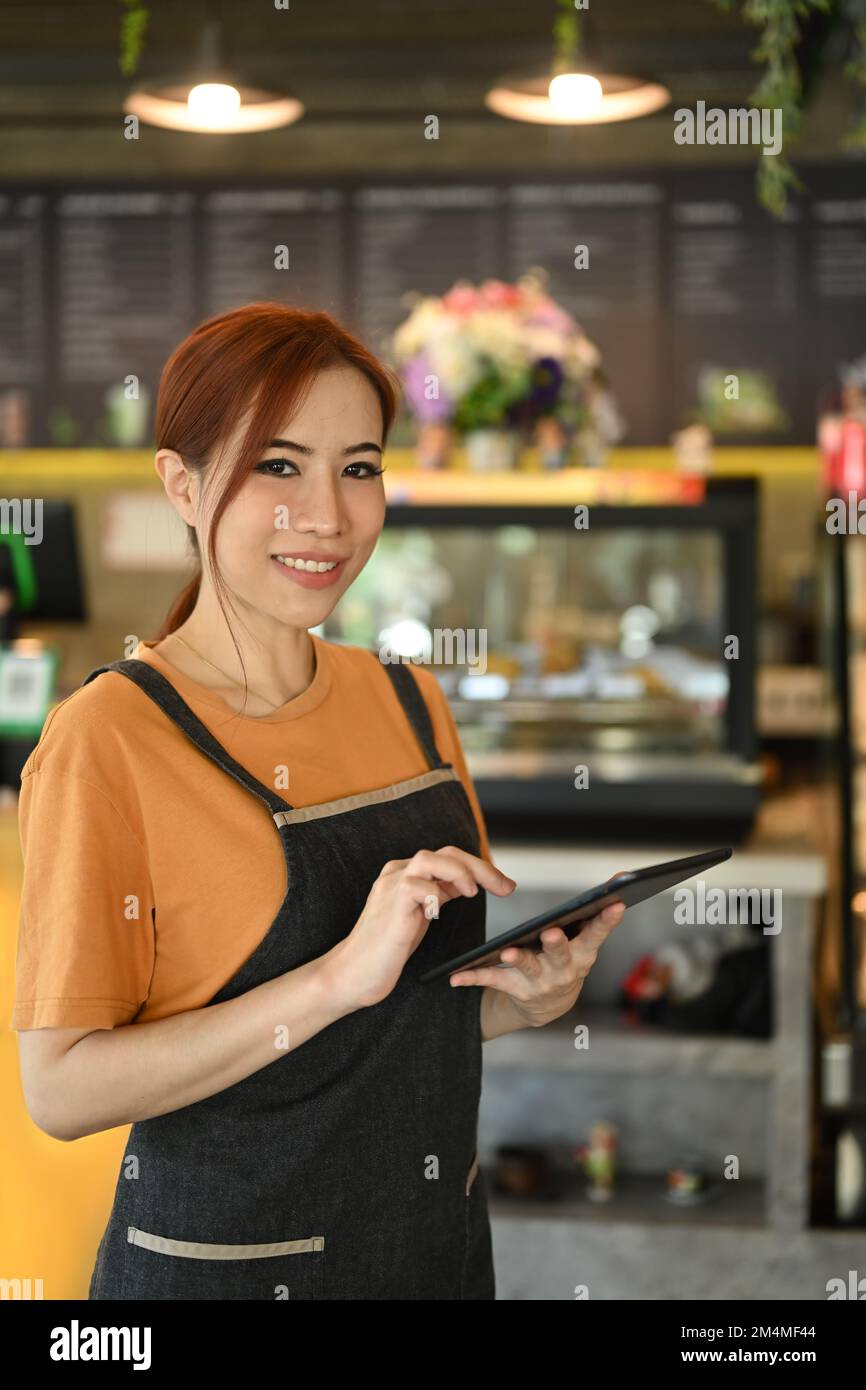 Portrait of smiling female entrepreneur wearing apron standing in front of counter of modern ...
