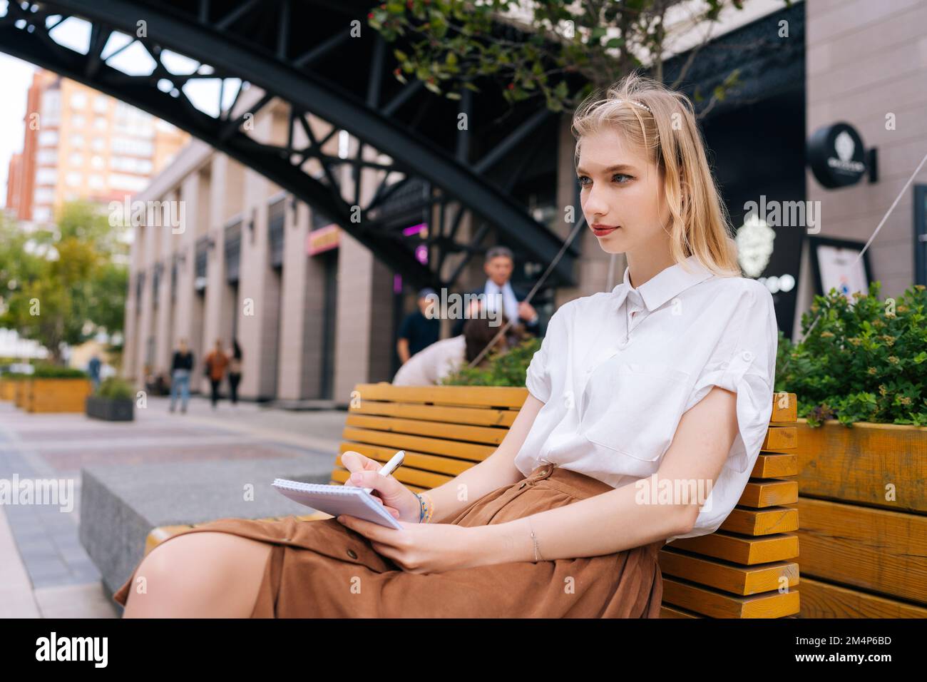 Pensive blonde young woman resting on bench in downtown writing plans ...