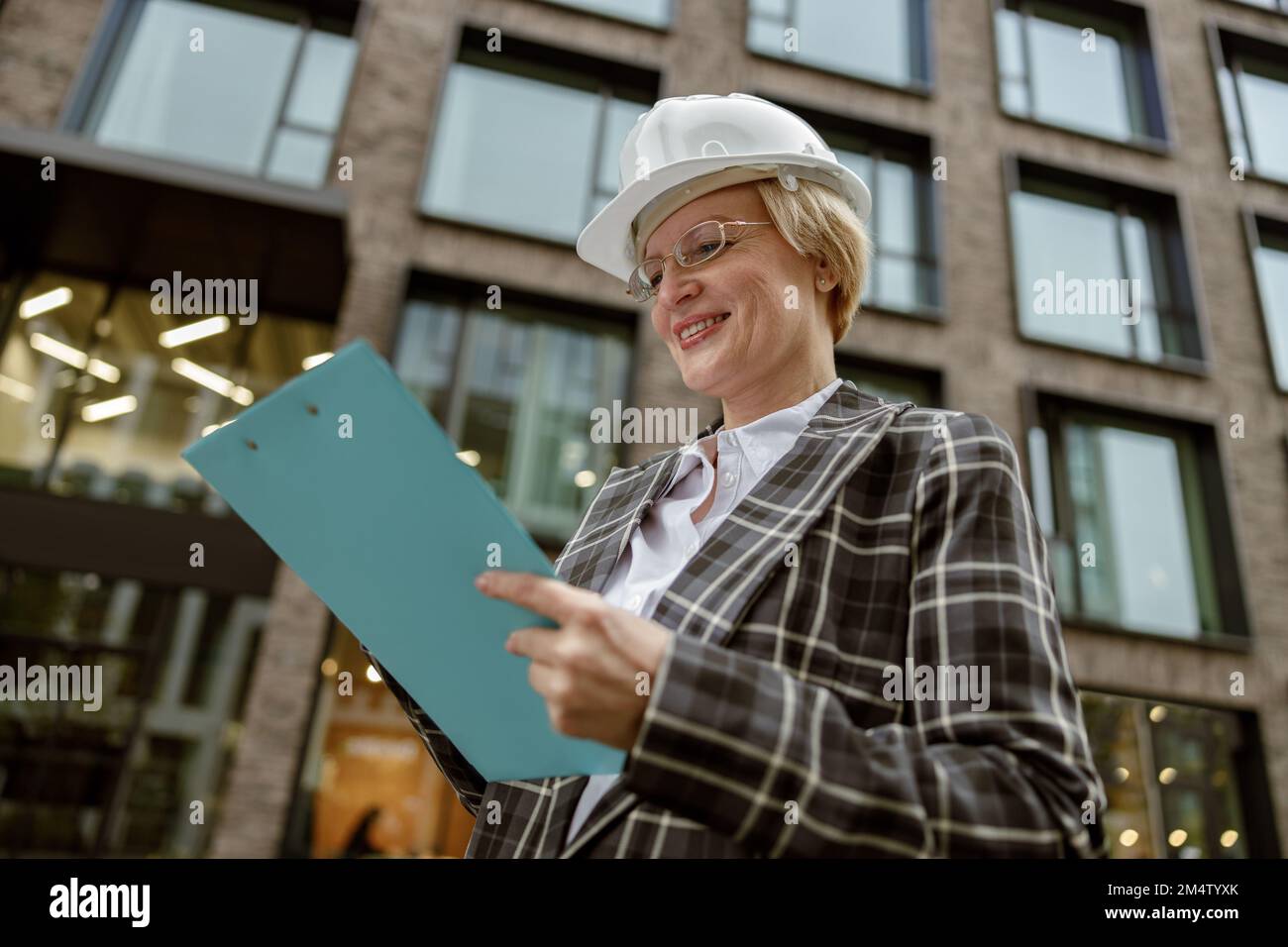 Senior female engineer in suit with white helmet making notes in tablet ...