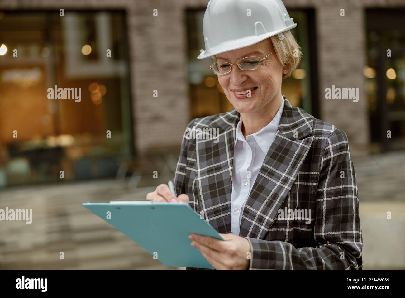 Smiling female engineer in suit with white helmet making notes in ...