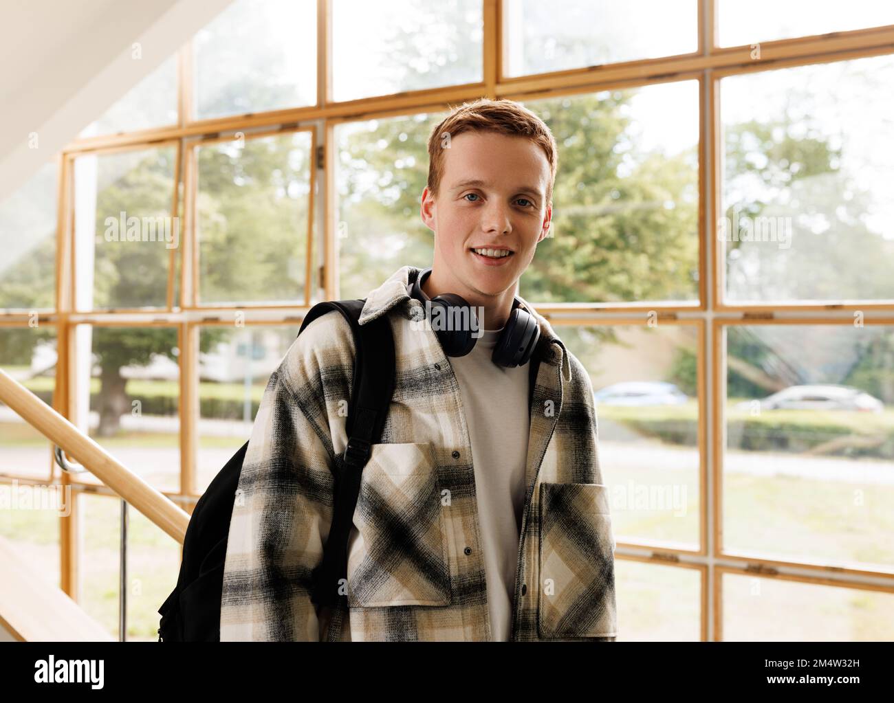 Portrait of a smiling male student standing against a window in college ...