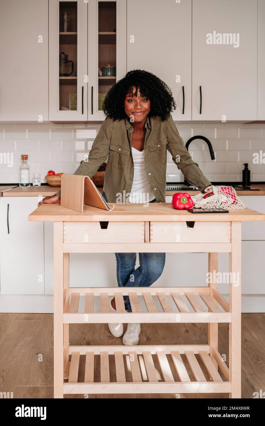 Smiling woman with tablet PC and mesh bag on kitchen island at home ...