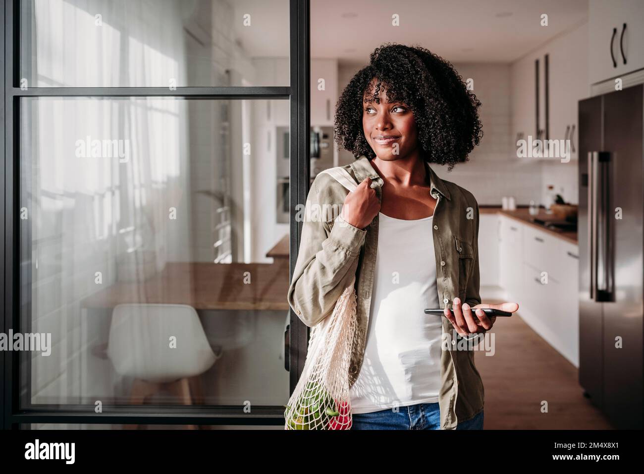 Thoughtful woman with mesh bag and smart phone standing at home Stock ...