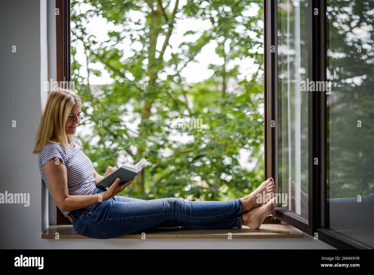 Woman reading book sitting on window sill at home Stock Photo - Alamy