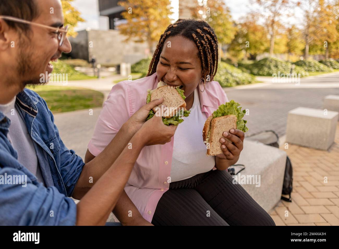 Happy man sharing sandwich with woman sitting on seat at footpath Stock ...