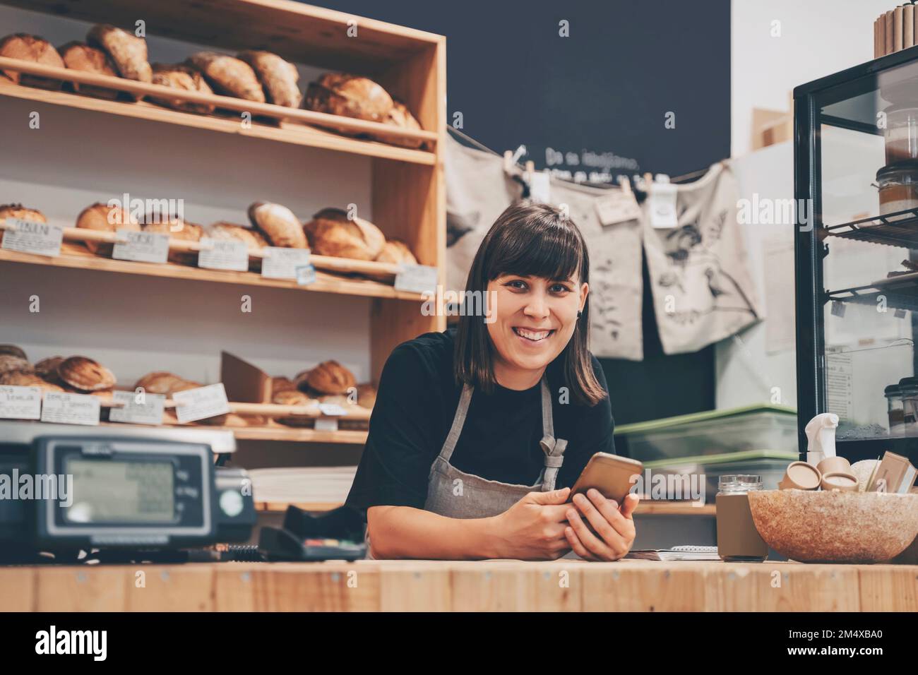 Smiling store owner with mobile phone leaning on checkout counter in ...