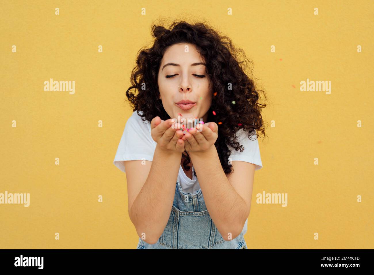 Woman blowing confetti in front of yellow wall Stock Photo - Alamy