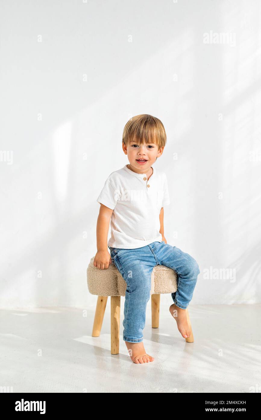 Cute boy sitting on stool in front of wall at home Stock Photo - Alamy