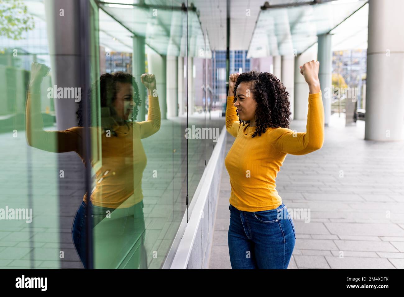 Woman flexing muscles looking reflection on glass wall Stock Photo - Alamy