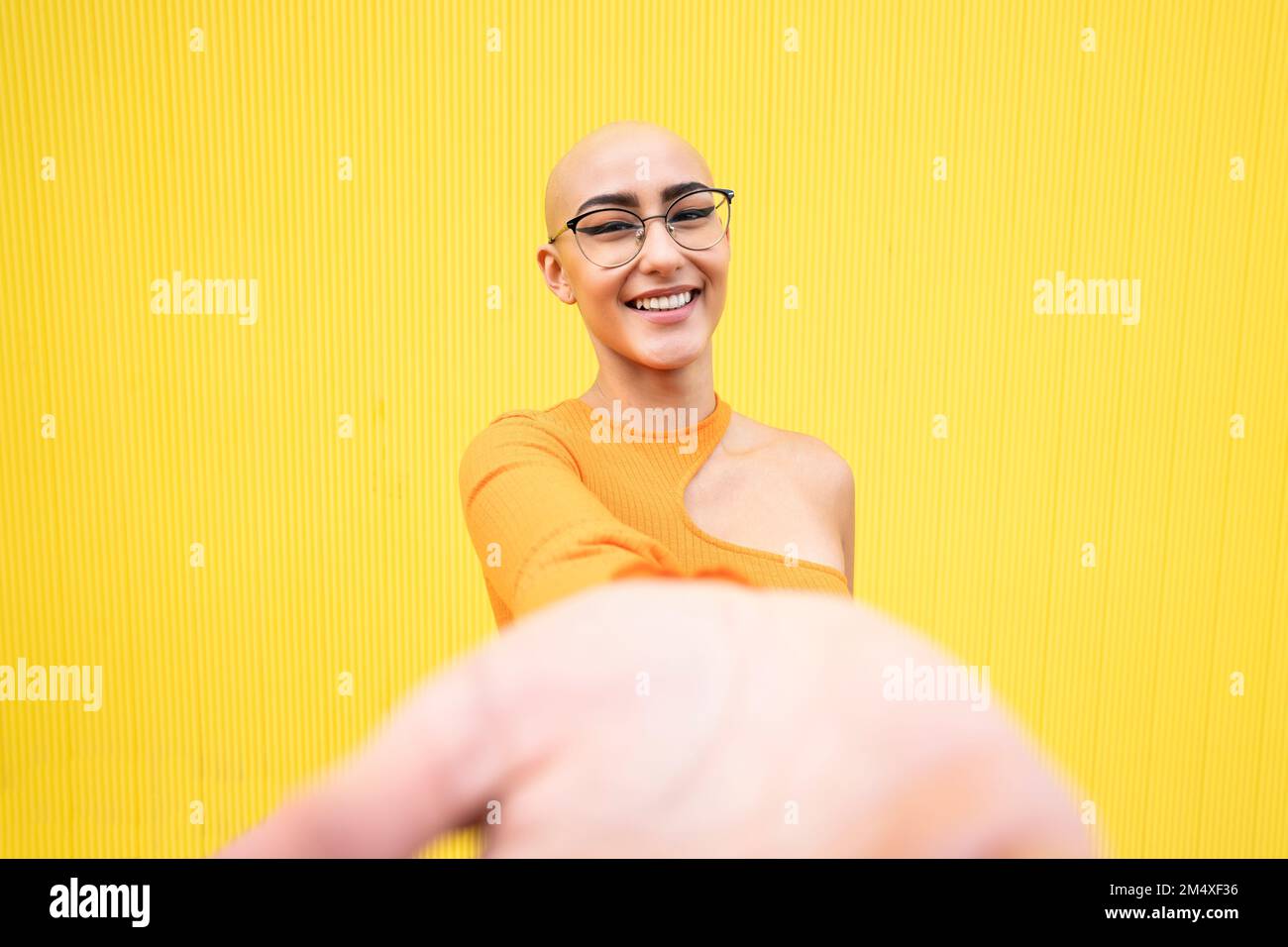 Happy woman with shaved head wearing eyeglasses in front of yellow wall ...