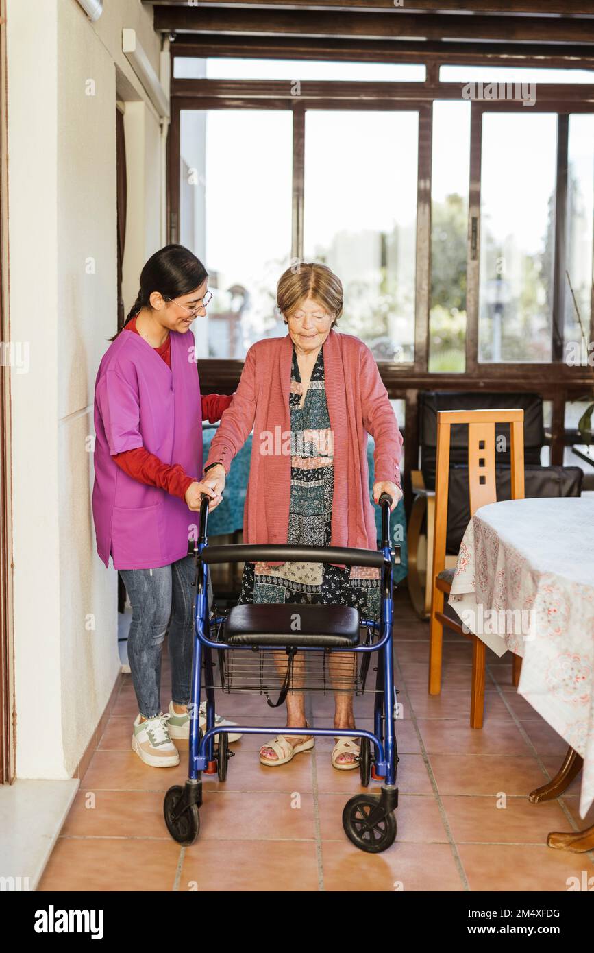 Smiling young caregiver helping senior woman to walk with mobility ...