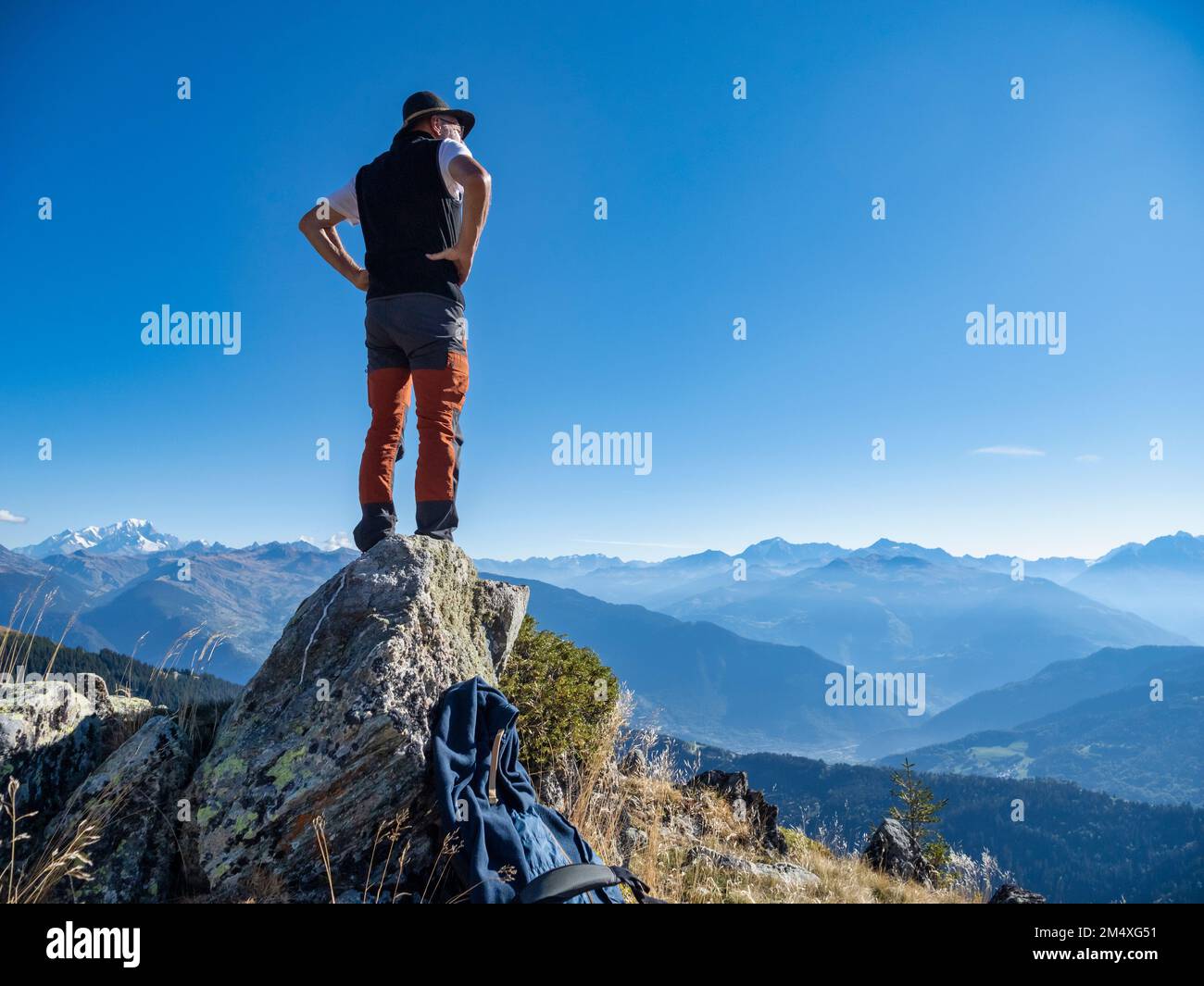 Senior man with hands on hip standing on rock under blue sky at Vanoise ...