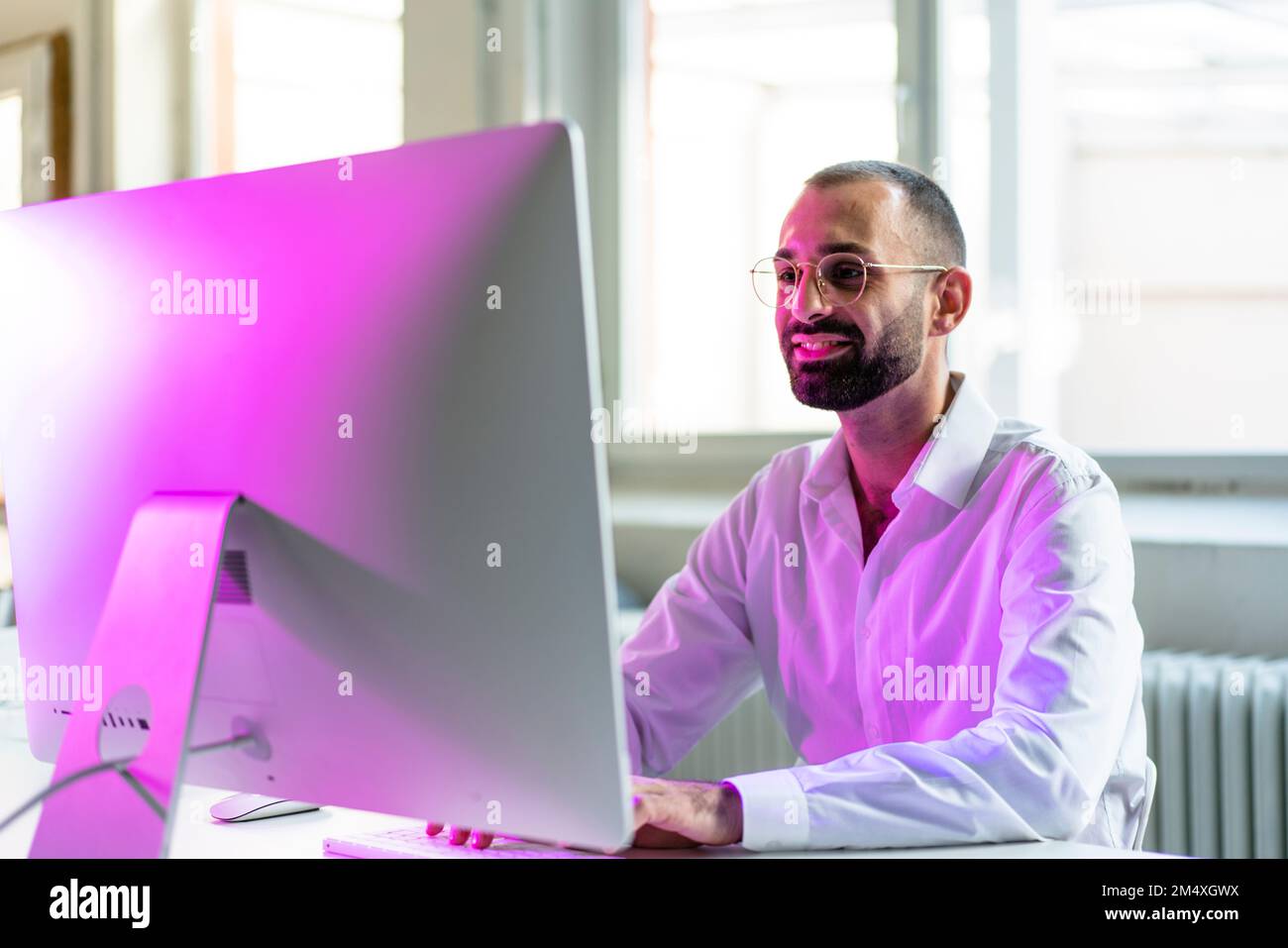 Pink light falling on smiling businessman using desktop PC at desk in ...