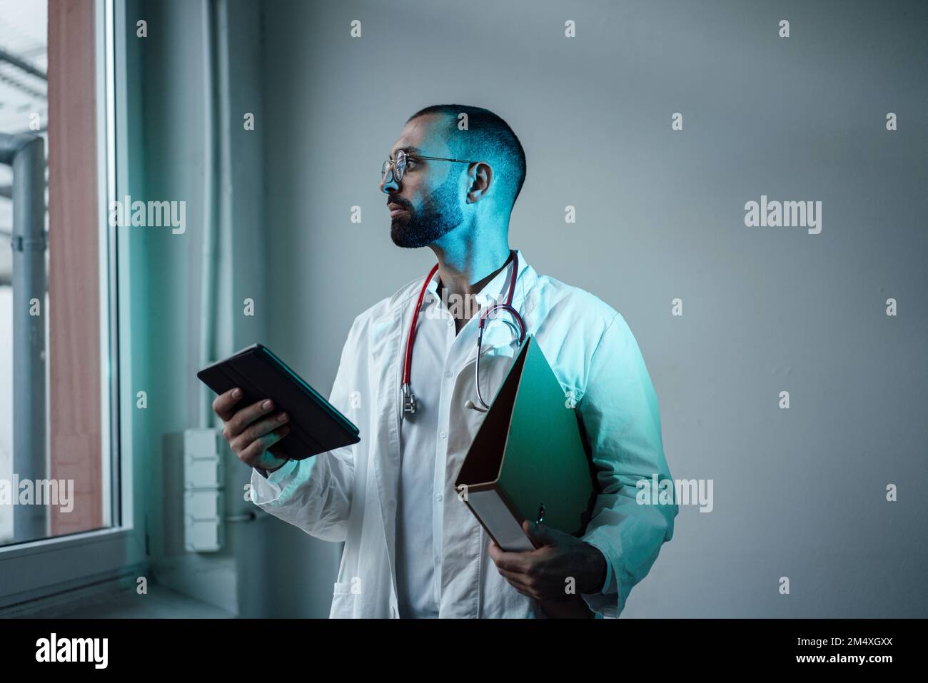 Thoughtful doctor holding tablet PC and file folder looking through ...