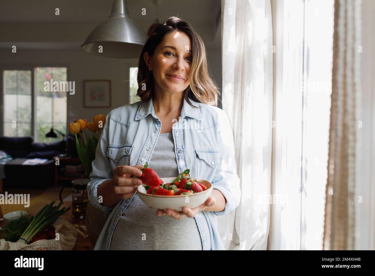 Smiling pregnant woman with bowl of strawberries standing at home Stock ...