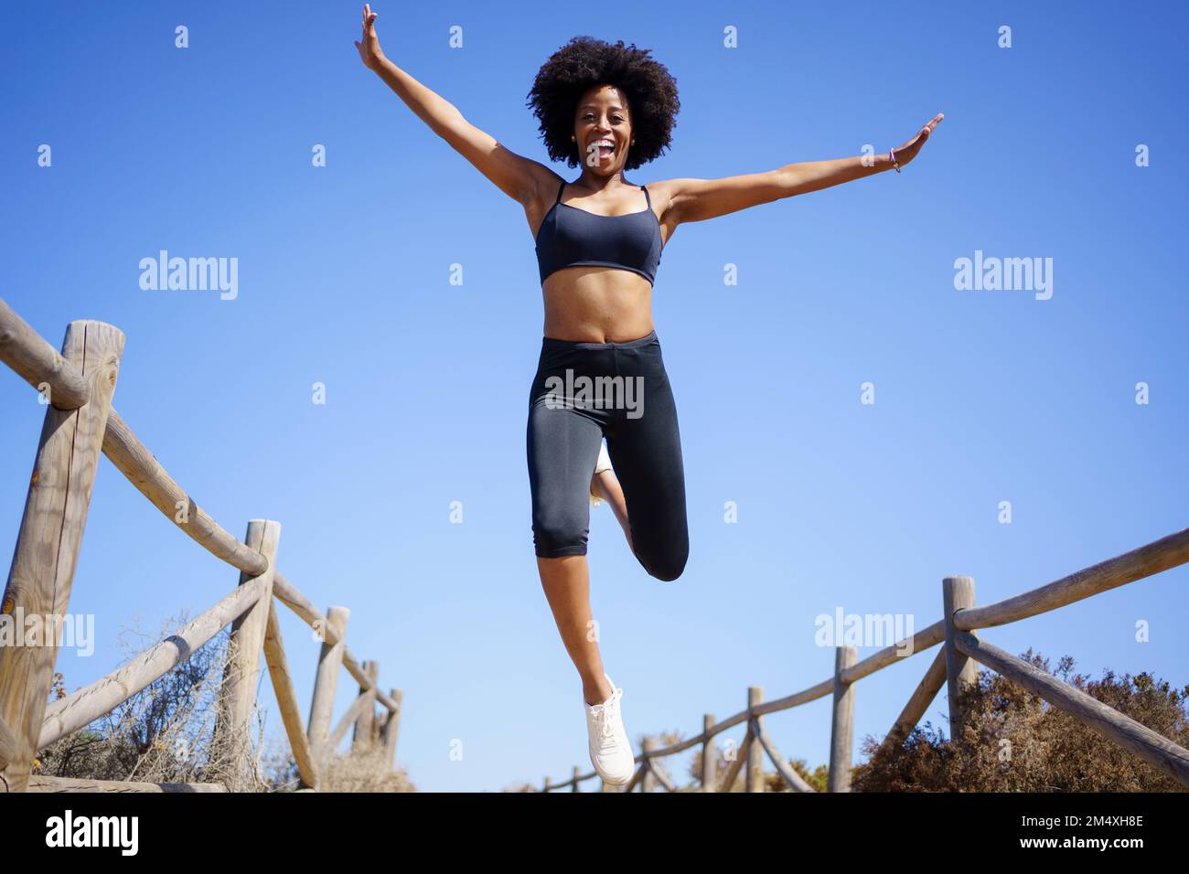 Cheerful young woman jumping over boardwalk under blue sky Stock Photo ...