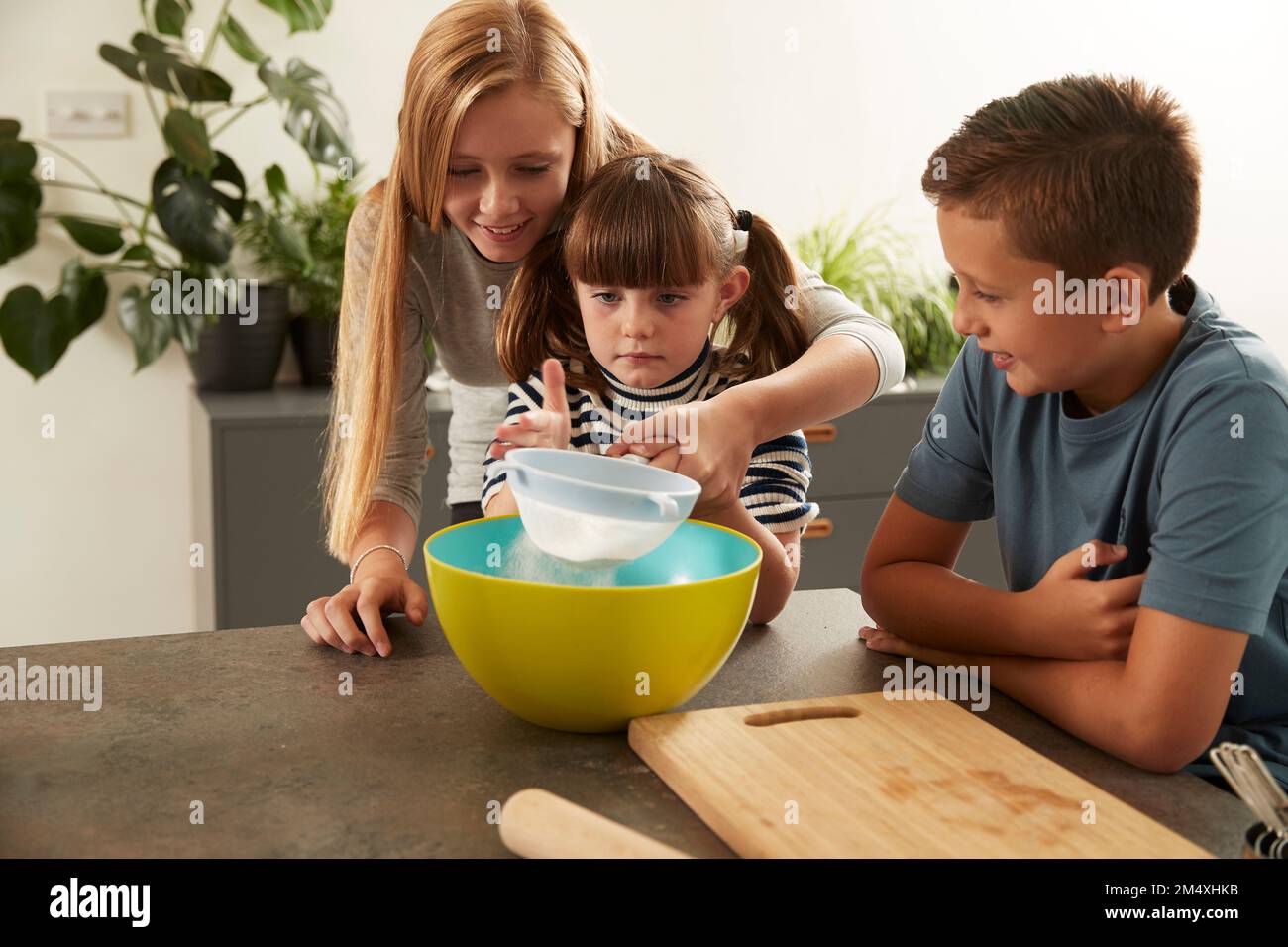 Smiling girl helping sister in sieving flour by brother at home Stock ...