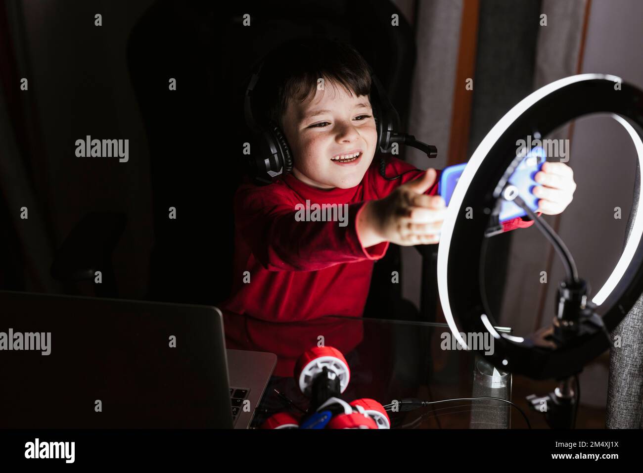 Happy boy sitting with laptop adjusting mobile phone on illuminated ...