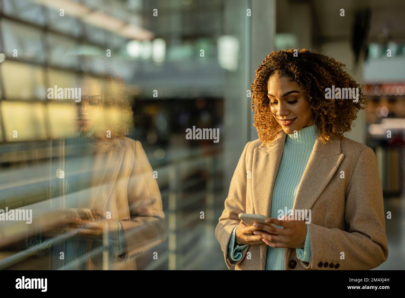 Smiling woman using smart phone by window with reflection on glass at ...