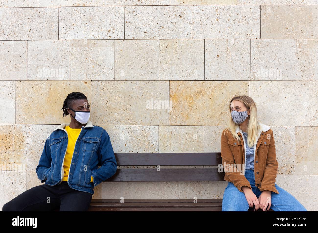 Young couple wearing protective face masks sitting on bench Stock Photo ...