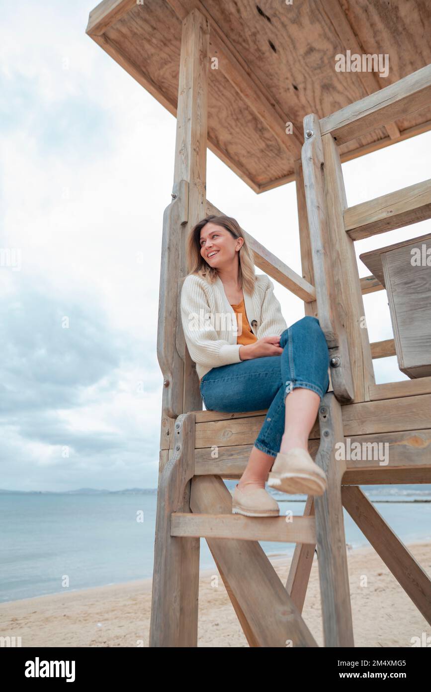 Smiling young woman sitting with legs crossed on wooden lifeguard hut ...