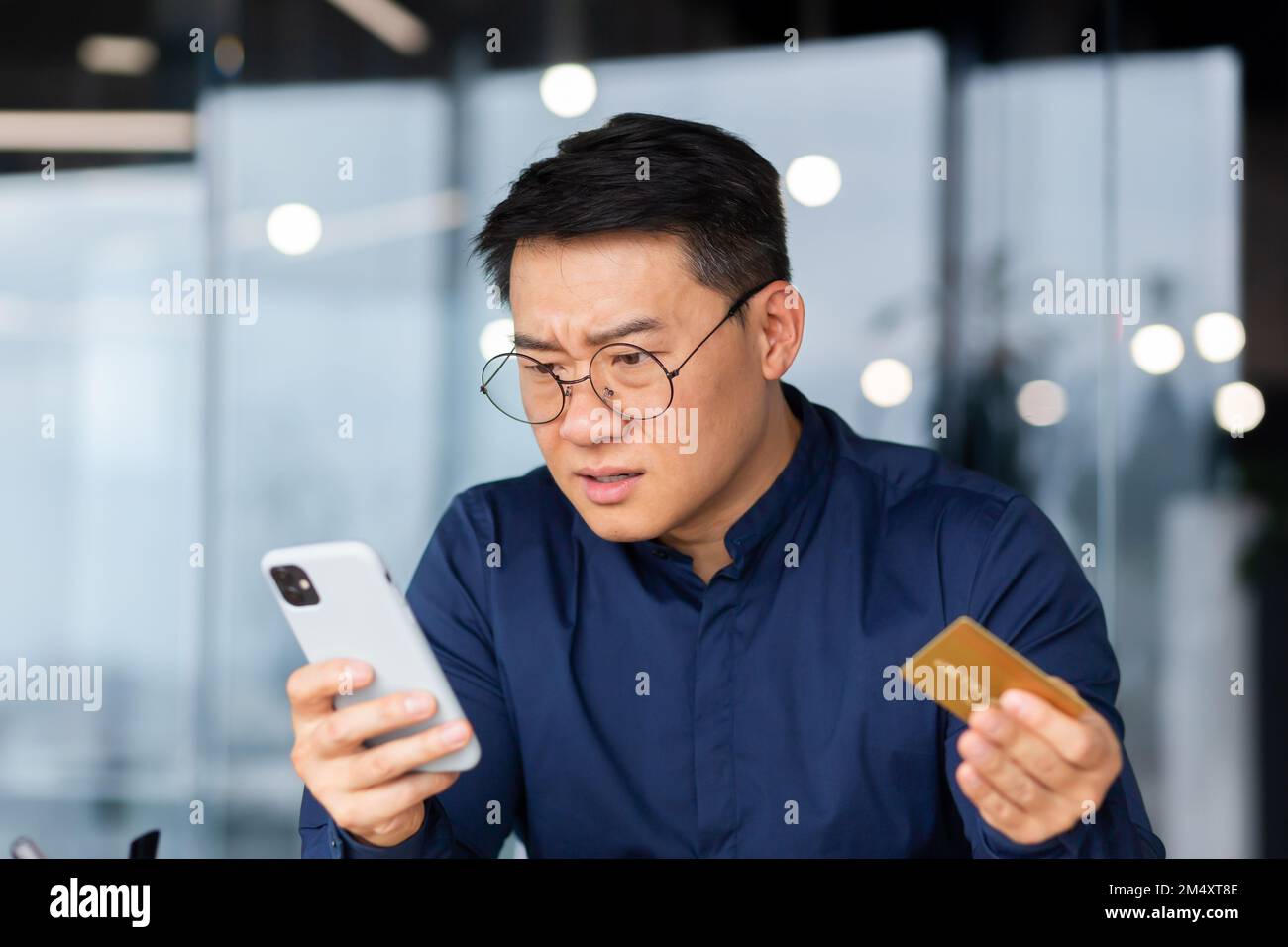 Close-up photo. A worried young Asian man in glasses holds a credit ...