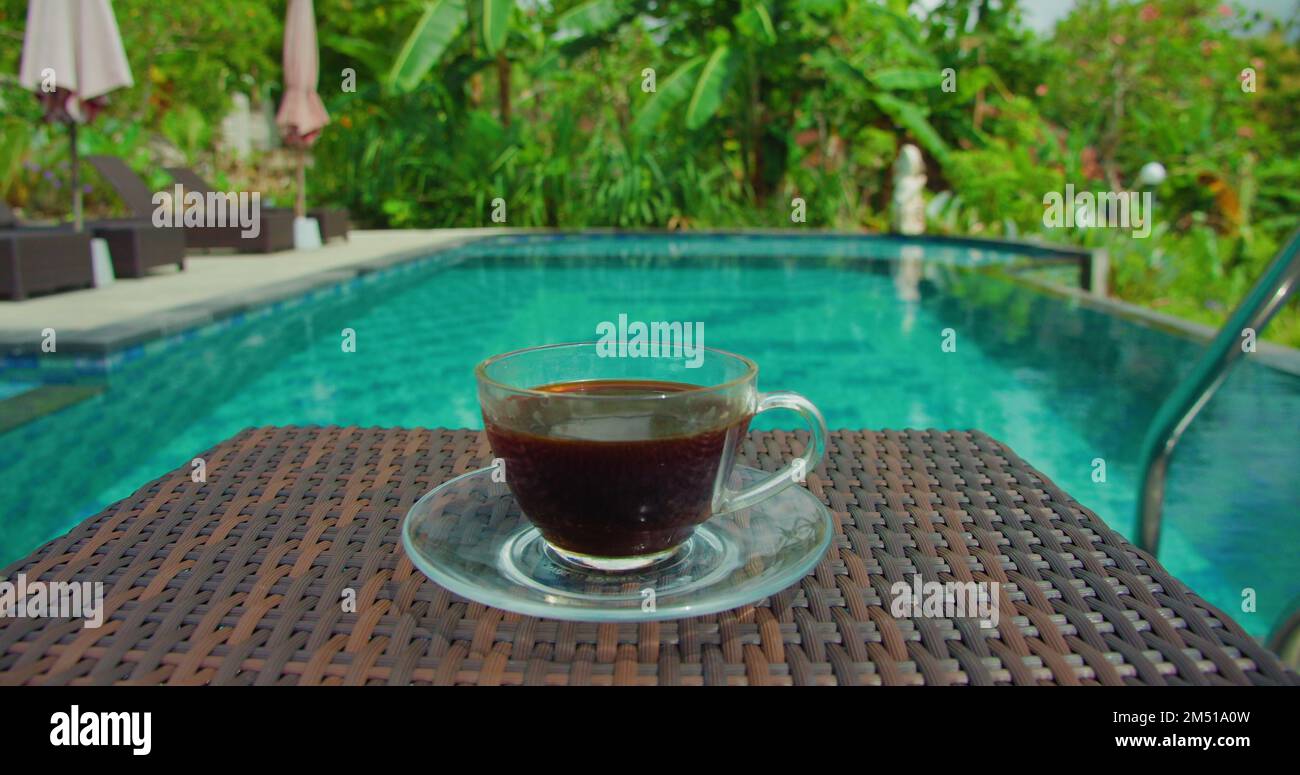 Cup of coffee served by blue water swimming pool in Bali island. Green ...