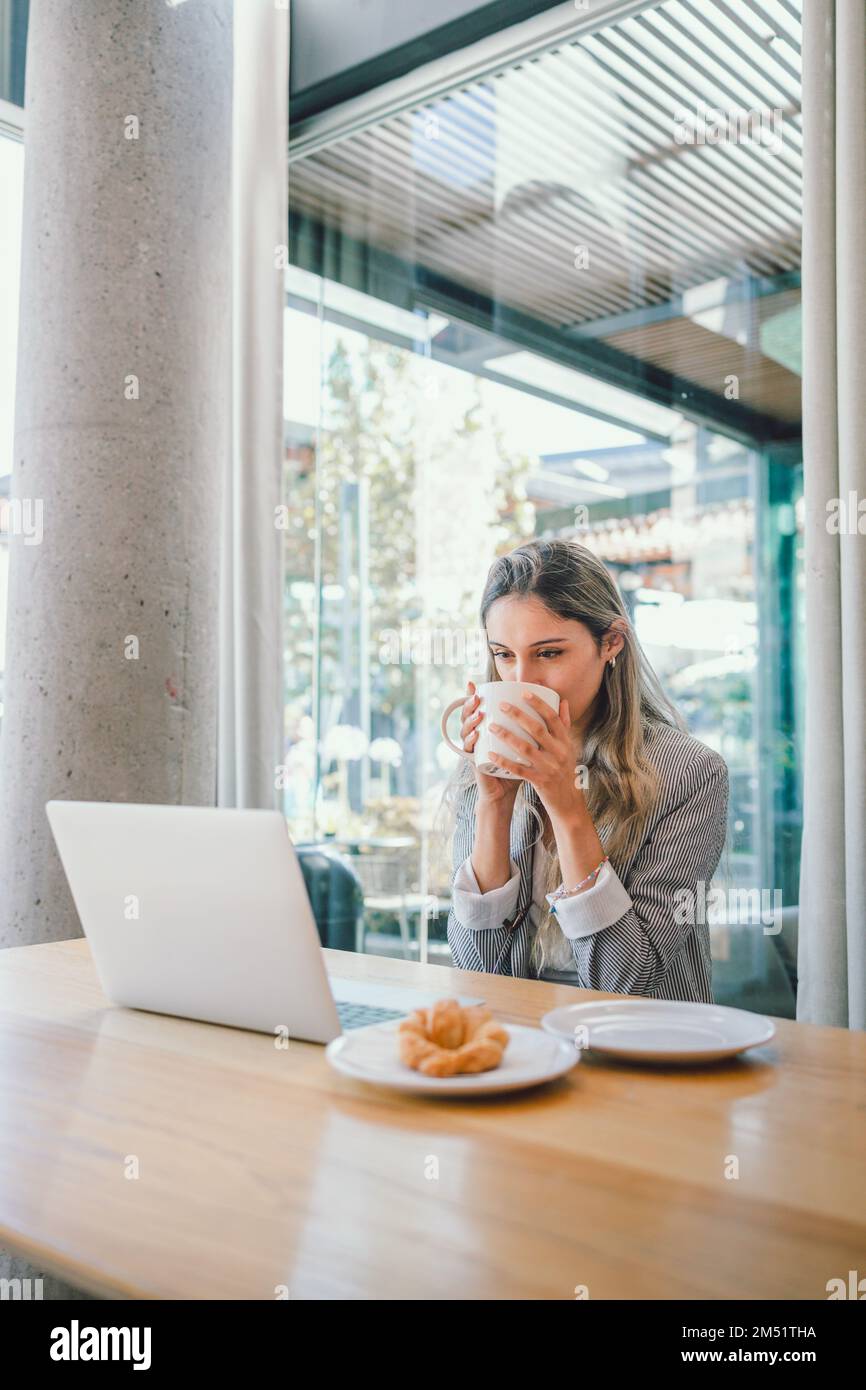 Young happy business woman having video call, drinking coffee from a mug in modern open ...