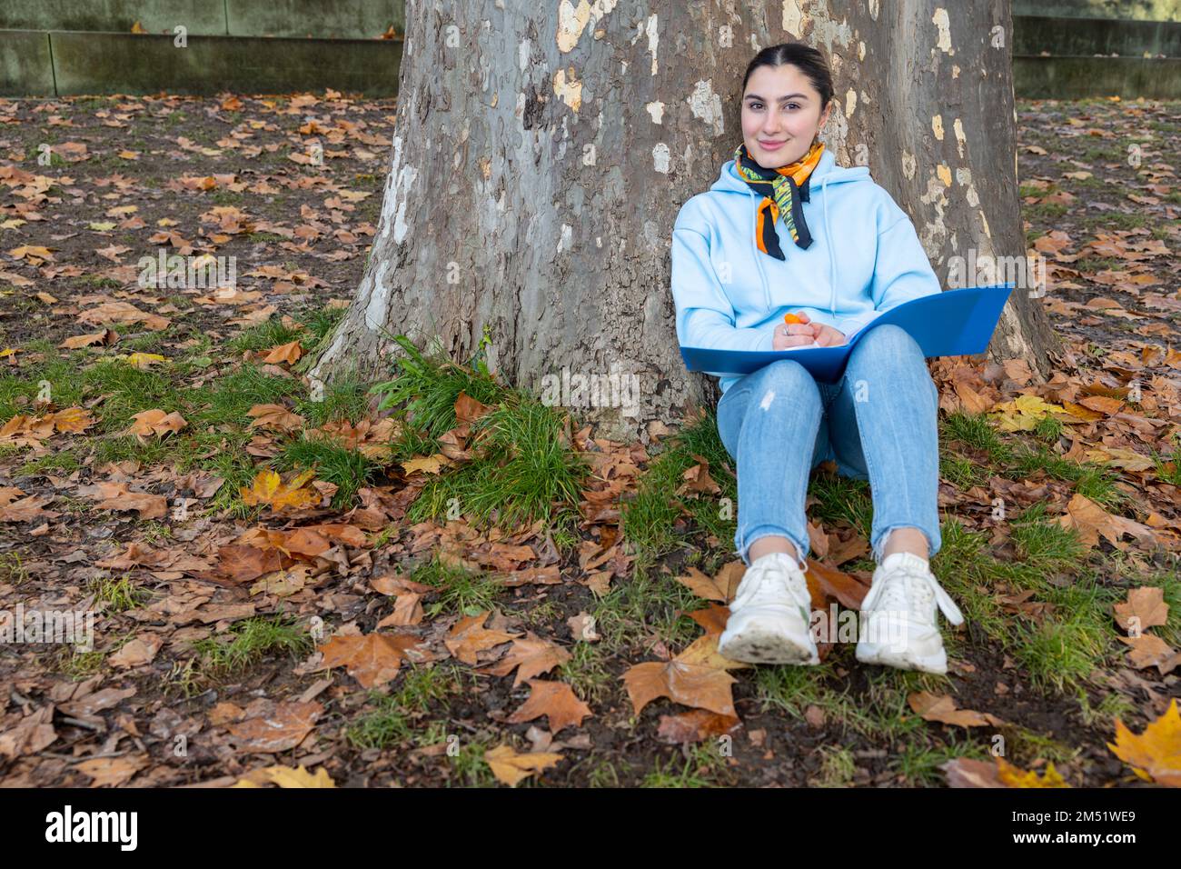 Oriental girl sitting under a tree with a blue folder in her hands ...