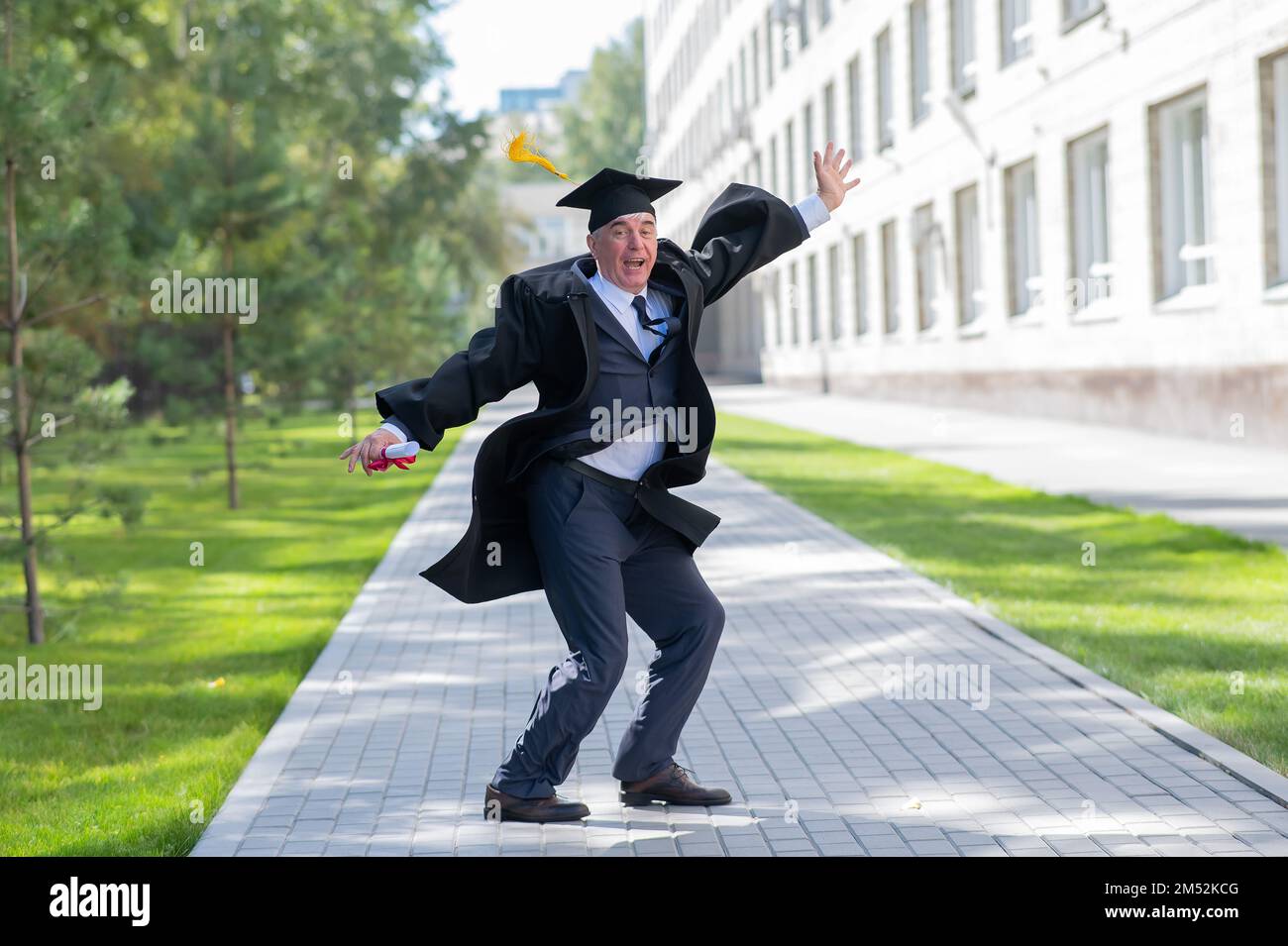 Old happy man in graduation gown jumping outdoors and holding diploma ...