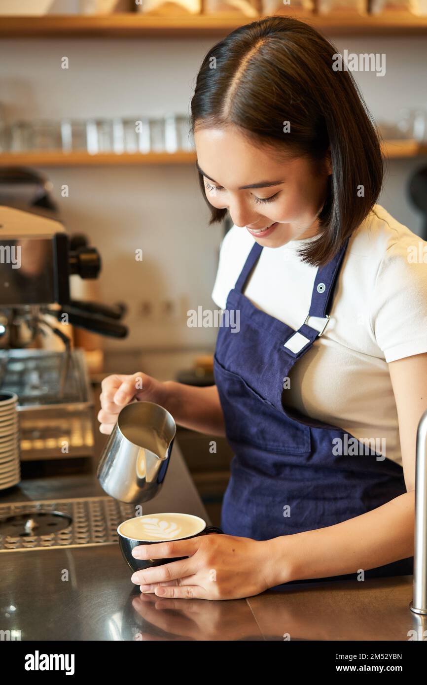 Smiling asian girl barista, cafe staff pouring steamed milk in coffee, prepare cappuccino with ...
