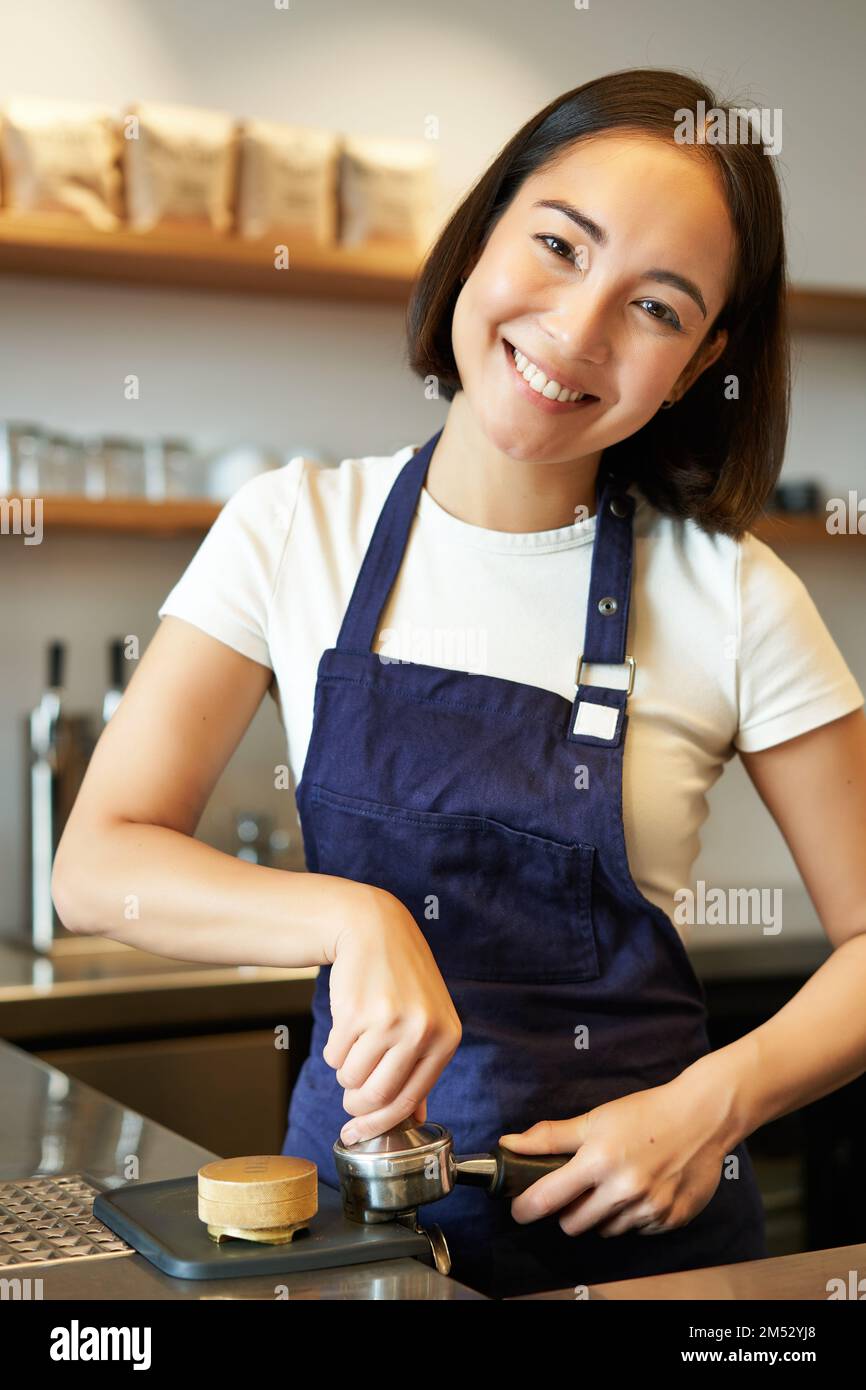 Vertical shot of cute asian barista, smiling girl puts coffee in tamper ...