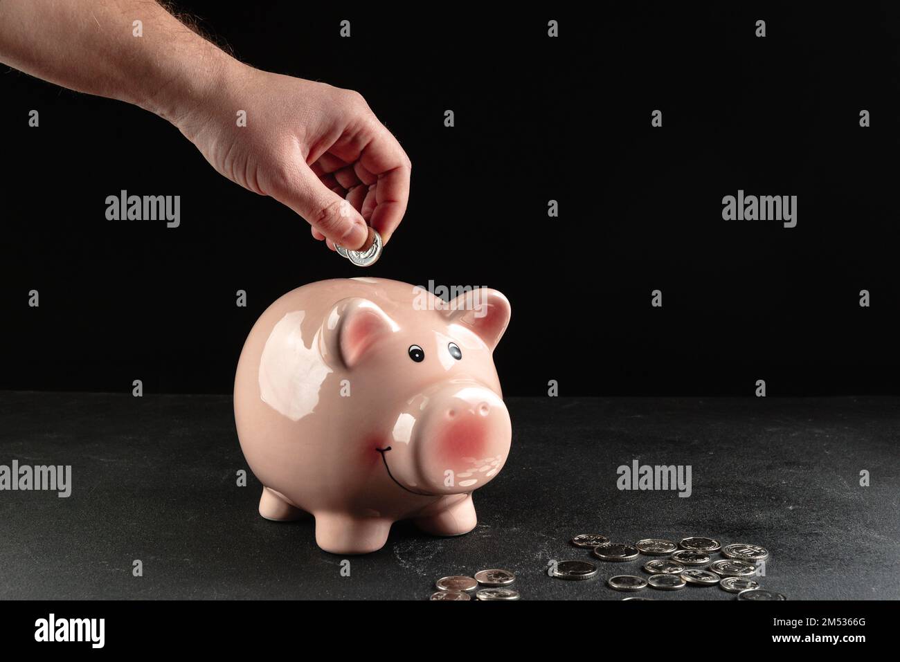 A person puts a coin in a piggy bank with a stack of coins next to it ...