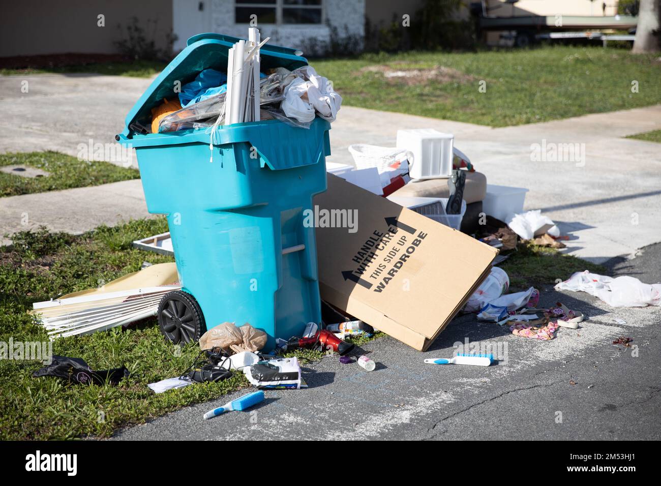 A blue plastic waste bin surrounded by overflowing trash on the side of ...