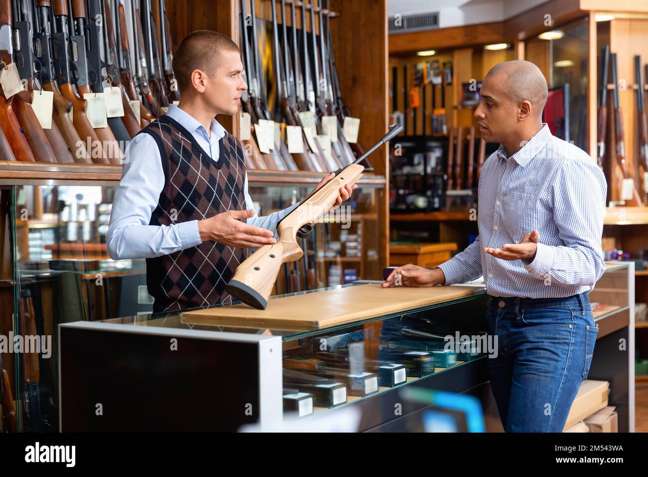 Salesperson helping Hispanic customer choose hunting rifle at gun store ...