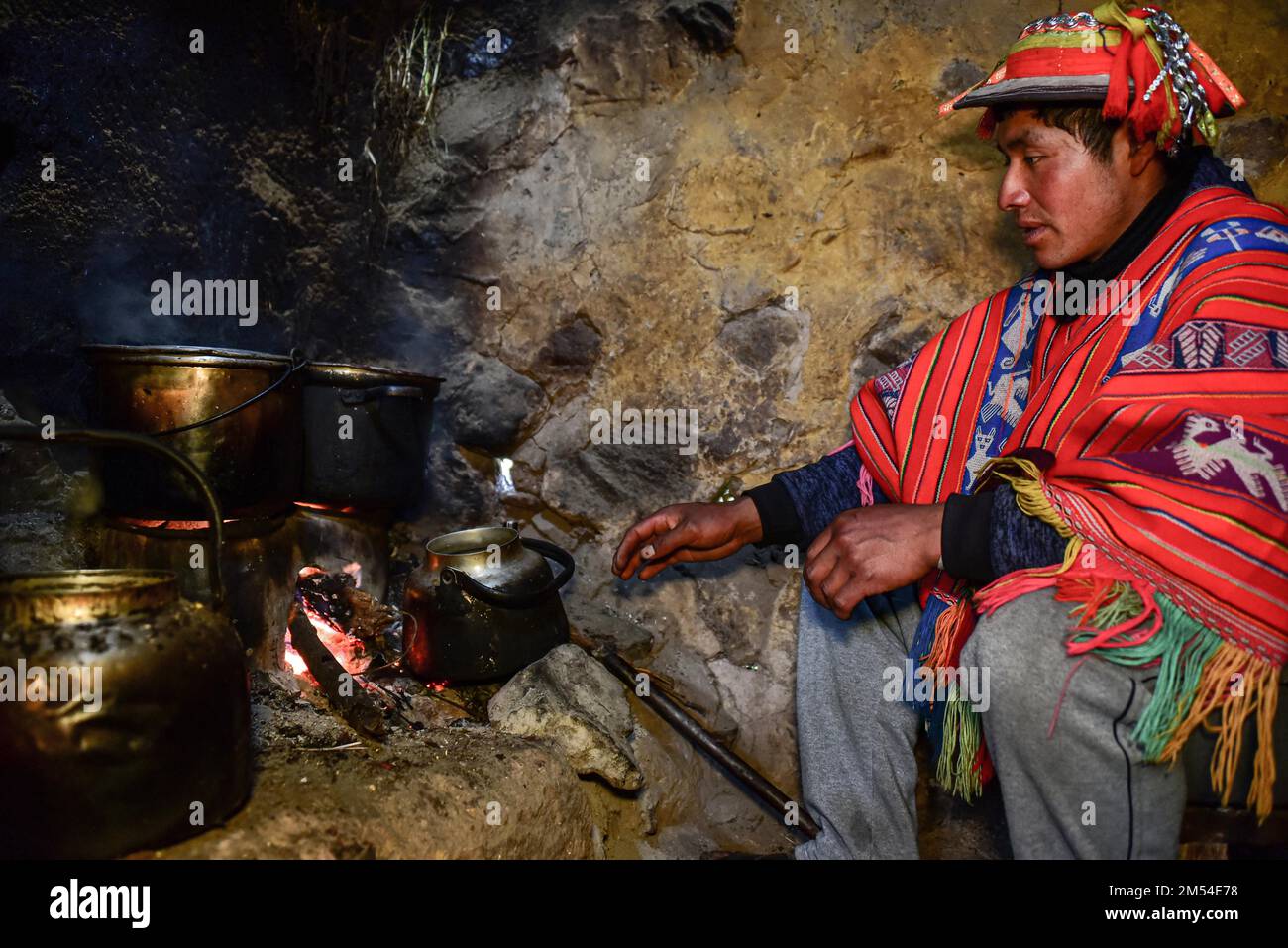 Quechua Indian in traditional dress cooking at the fireplace of his hut ...