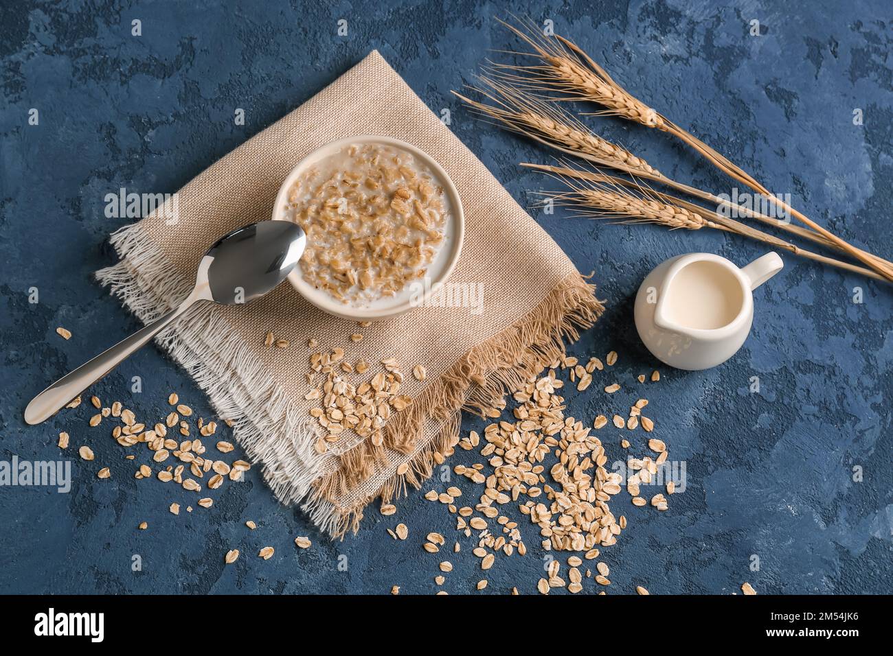 Bowl of tasty oatmeal, pitcher with milk, scattered flakes and wheat ...