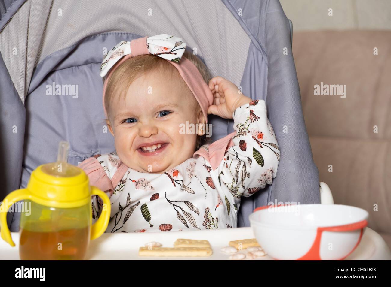A happy little girl sits in a high chair with a bottle and a plate and ...