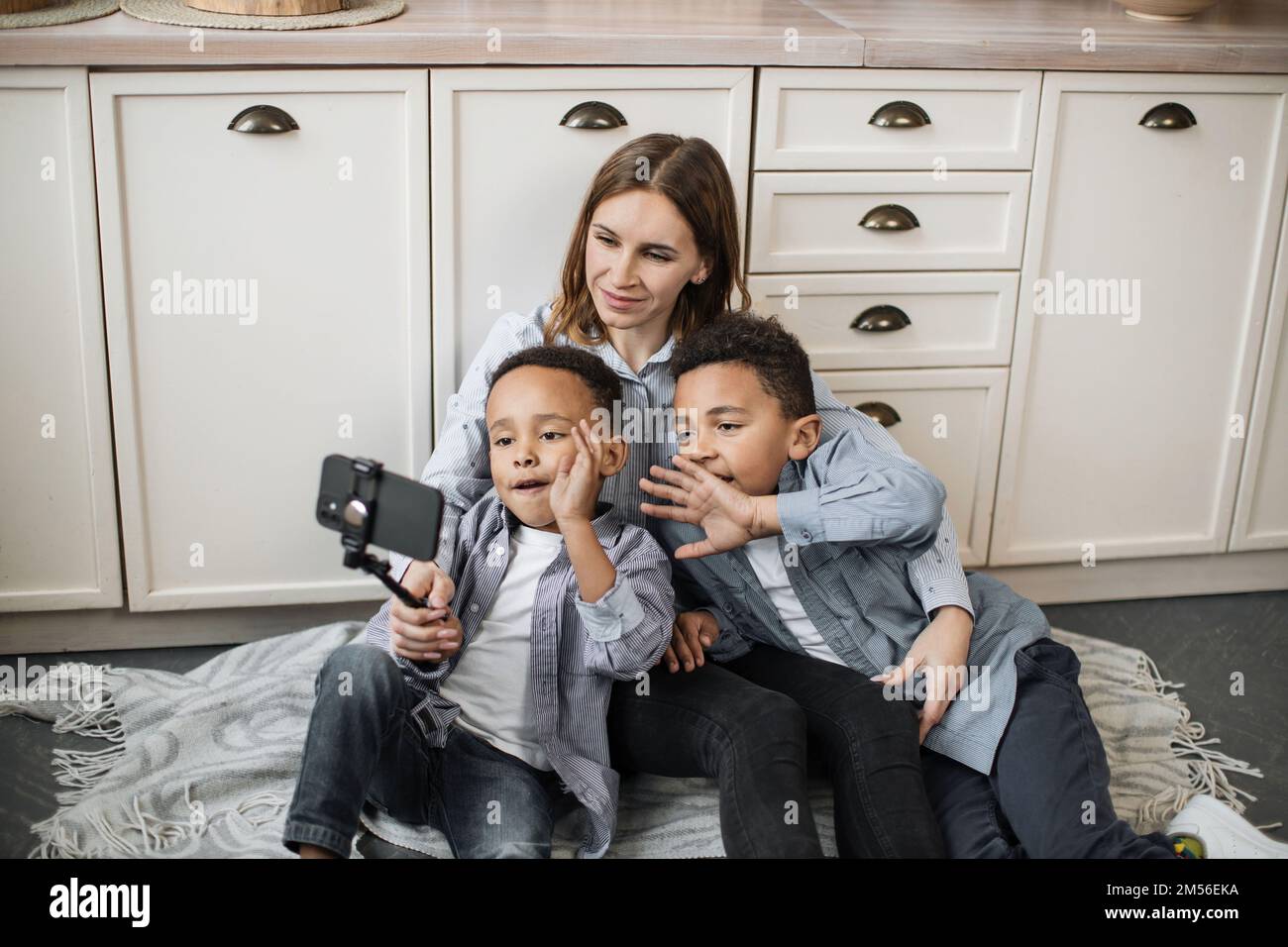 Happy multinational family of mother and two sons sitting on warm floor ...