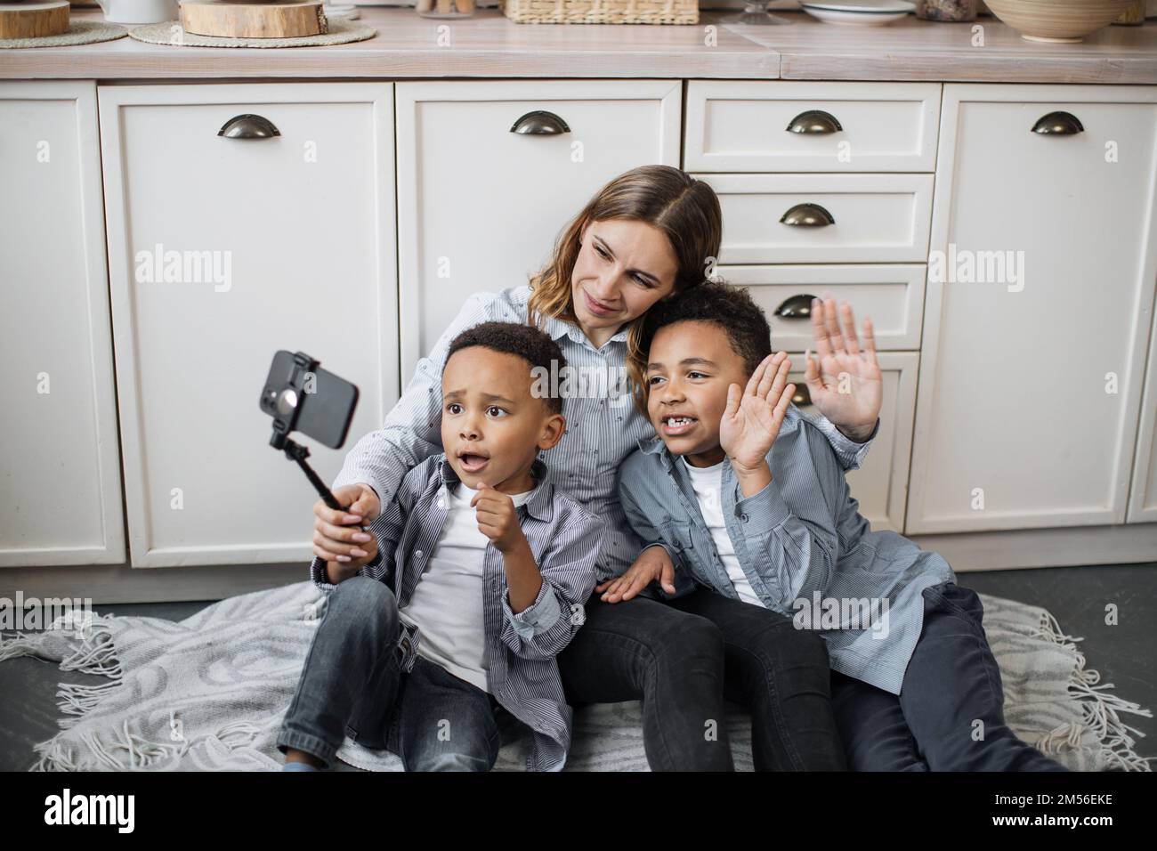 Happy multinational family of mother and two sons sitting on warm floor ...