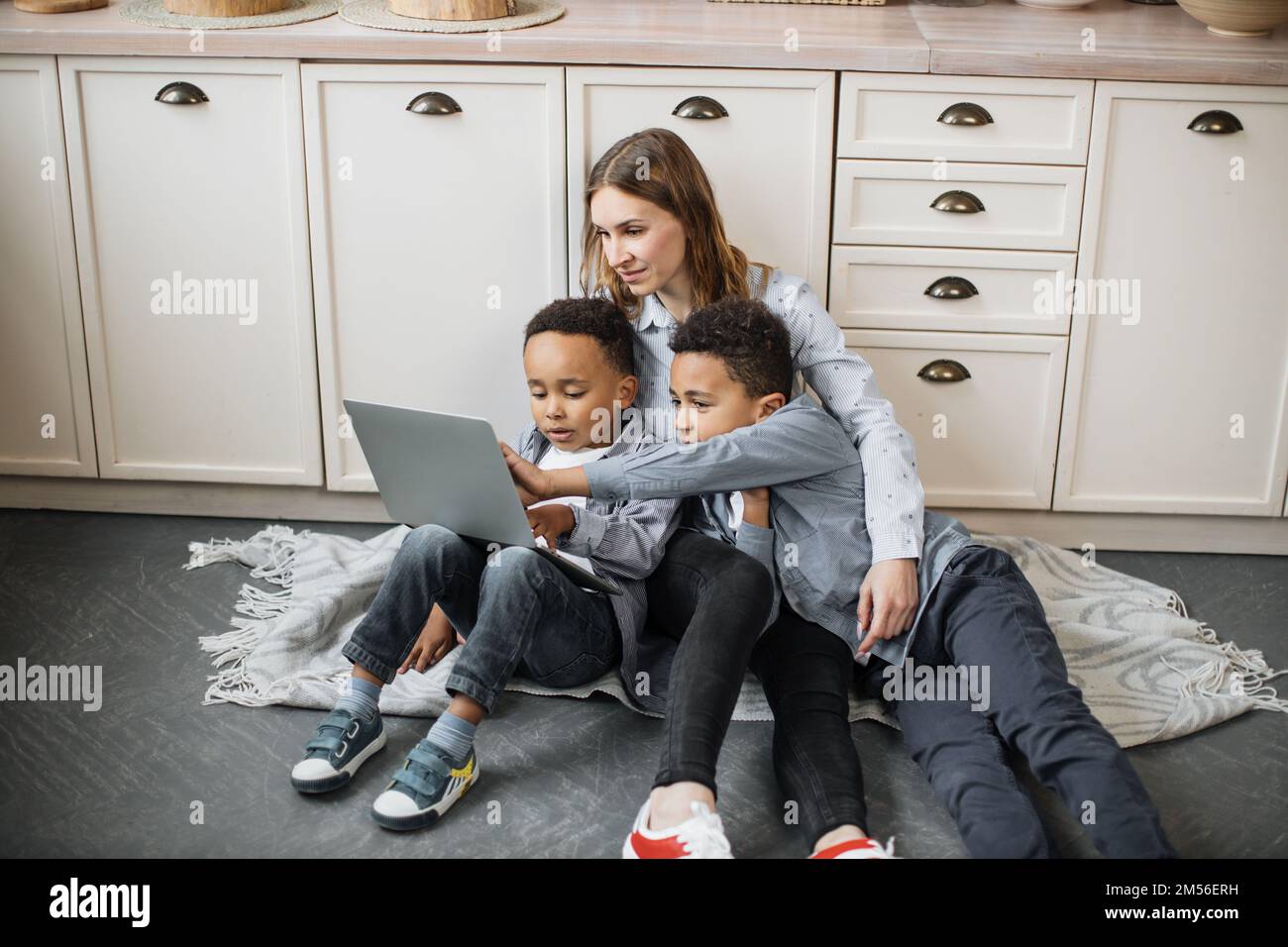Happy multinational family of mother and two sons sitting on warm floor ...