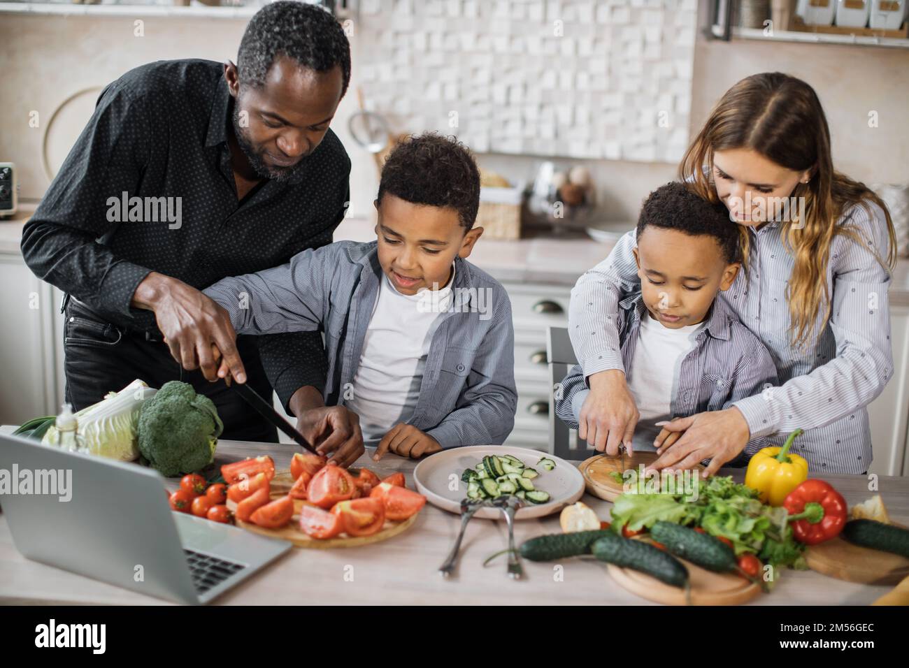 Happy young parents teaching little preschooler boys to chop vegetables ...