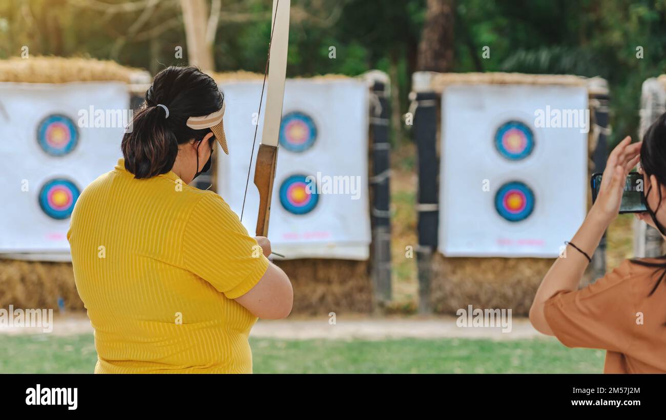 Back view of Asian woman wear visor with face mask aims archery bow and ...